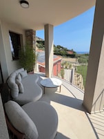 Uma varanda com sofá, mesa pequena, um pilar de pedra e vista para o mar e casas.
