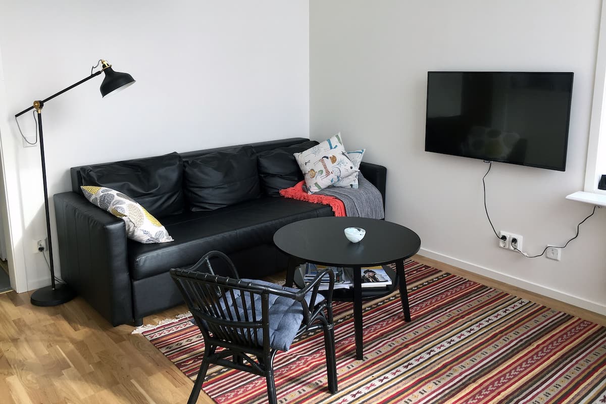 Uma sala de estar moderna com um sofá de couro preto, uma mesa de jantar preta, uma cadeira preta, um abajur e uma TV de tela plana fixada na parede.