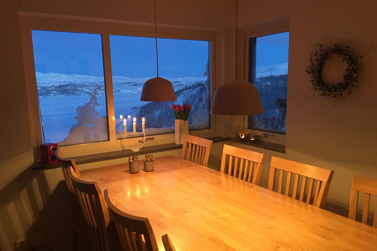 Uma área de refeições com mesa e cadeiras de madeira, um candelabro com velas acesas e vista para uma paisagem nevada através de grandes janelas.