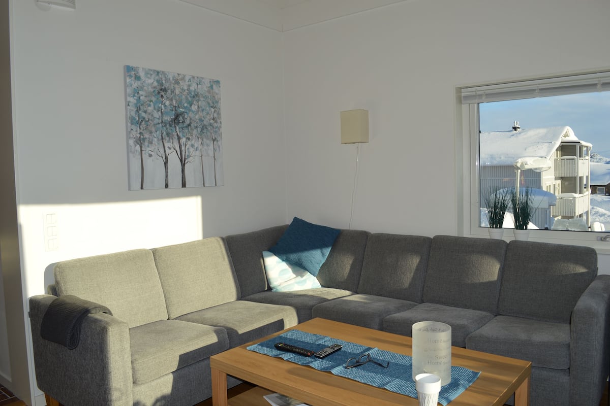 Uma sala de estar com um sofá cinzento, uma mesa de centro de madeira e uma pintura de árvores na parede.