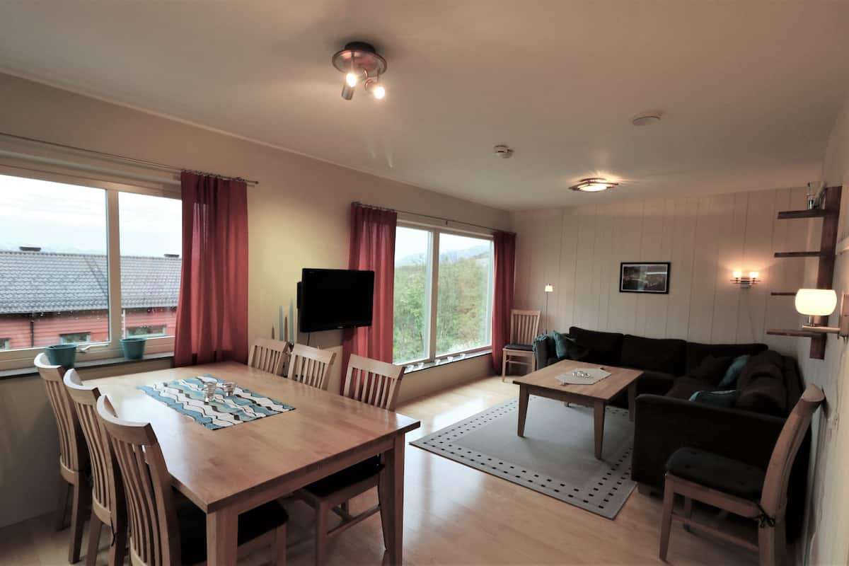 Uma sala de estar ampla com uma mesa de jantar de madeira, cadeiras, um sofá e uma TV. Grandes janelas com cortinas oferecem luz natural e vista para o exterior.