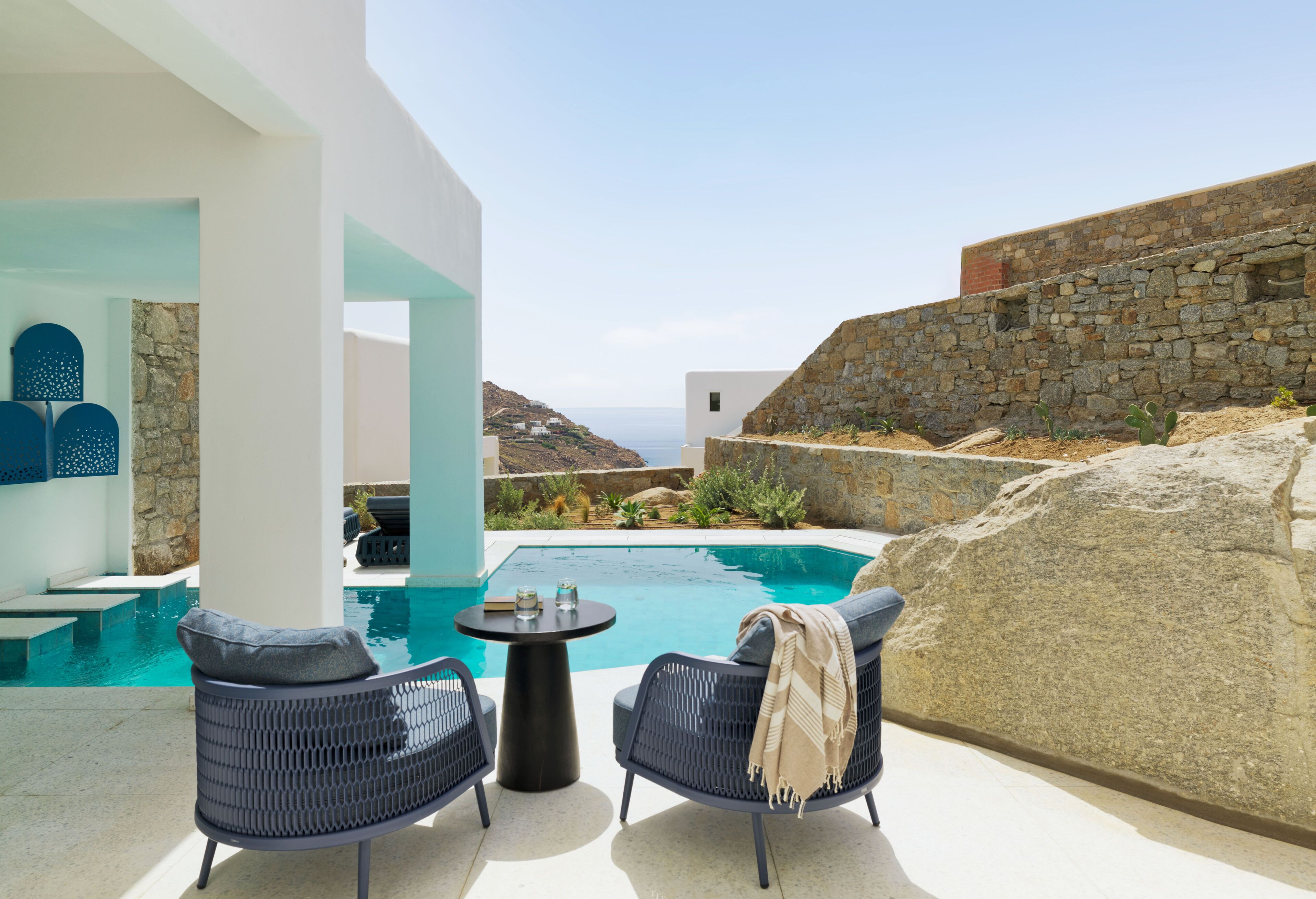 Uma área de piscina moderna ao ar livre, com duas cadeiras de vime e uma mesa pequena, com vista para uma paisagem deslumbrante do mar e colinas rochosas.