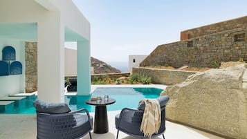 Uma área de piscina moderna ao ar livre, com duas cadeiras de vime e uma mesa pequena, com vista para uma paisagem deslumbrante do mar e colinas rochosas.