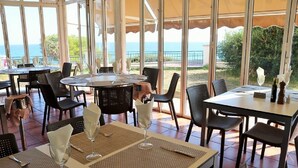 Restaurante com mesas ao ar livre, toalhas brancas e copos de vinho, e vista para o mar.