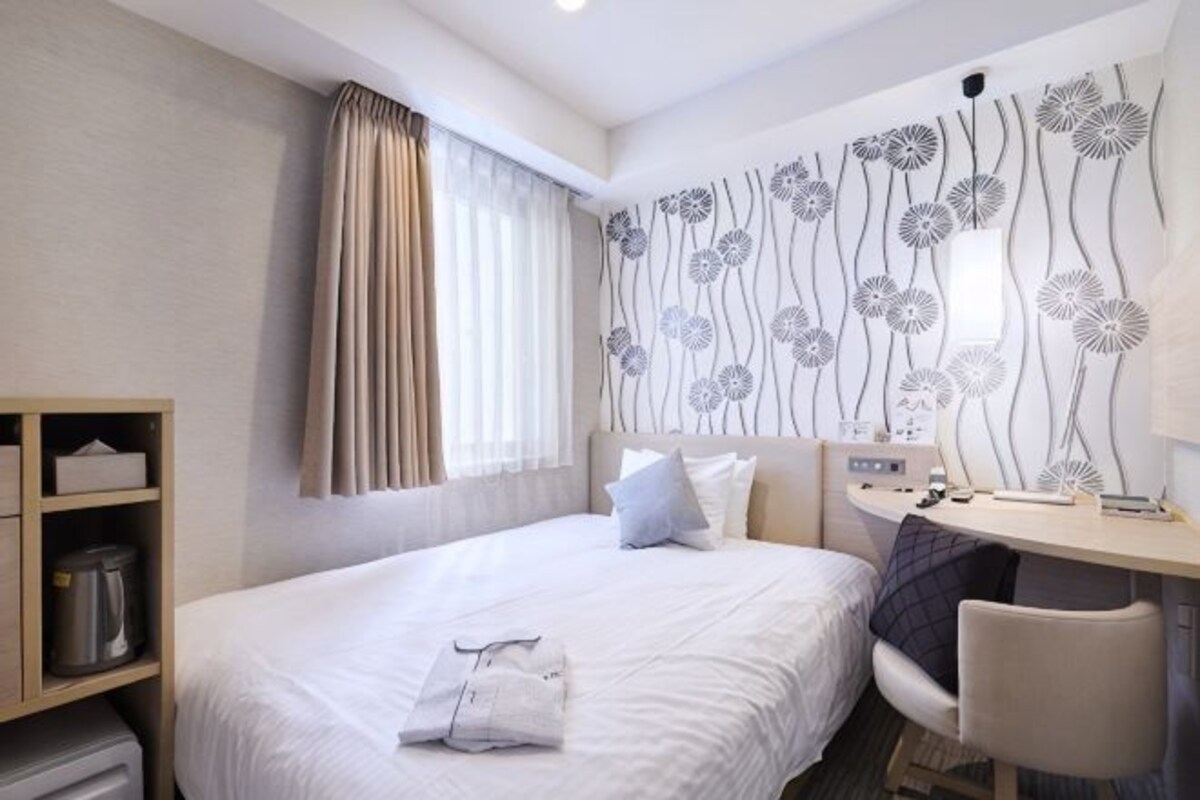 Quarto de hotel com cama, escrivaninha, cadeira, armário e janela com cortinas.