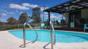 Piscina com escadas de metal, área de estar coberta com cadeiras e vista para montanhas ao fundo.