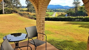 Pátio com mesa e cadeiras, com vista para área gramada, estrada e colinas ao longe.