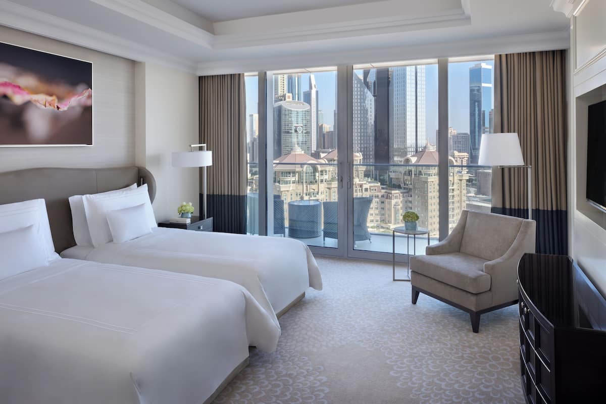 Quarto de hotel com duas camas, uma cadeira, uma televisão e uma janela grande com vista para a cidade.