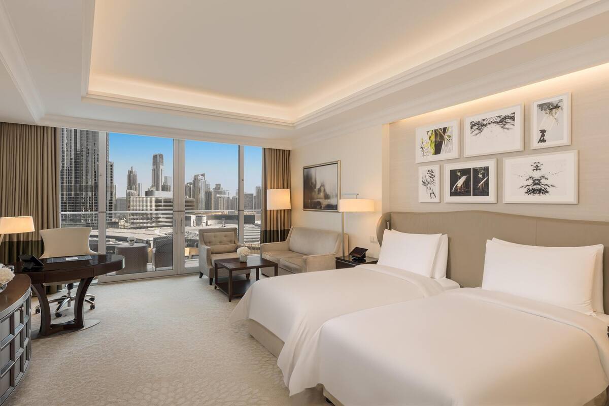 Quarto de hotel com duas camas, uma escrivaninha, uma cadeira, uma TV e uma janela grande com vista para a cidade.