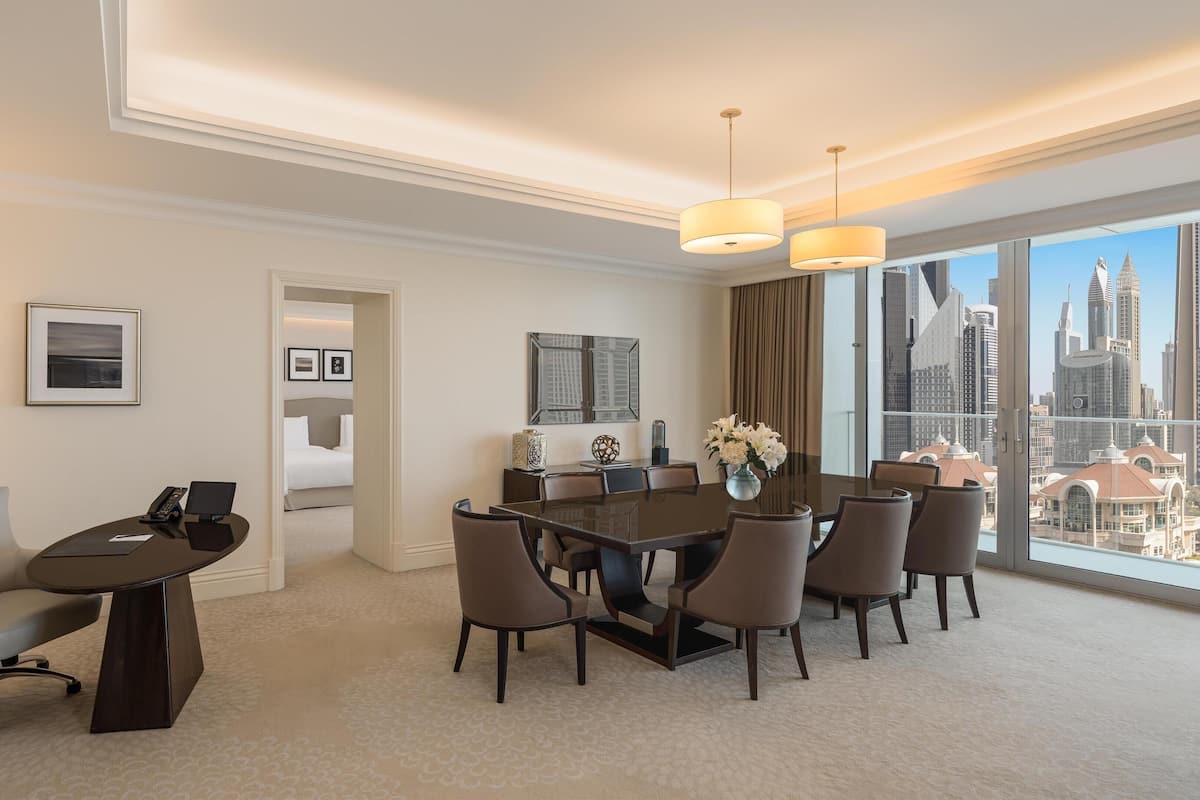 Quarto de hotel com uma grande mesa de jantar, seis cadeiras, uma escrivaninha com telefone, abajur, obras de arte emolduradas, um vaso com flores e vista para a cidade através de grandes janelas.