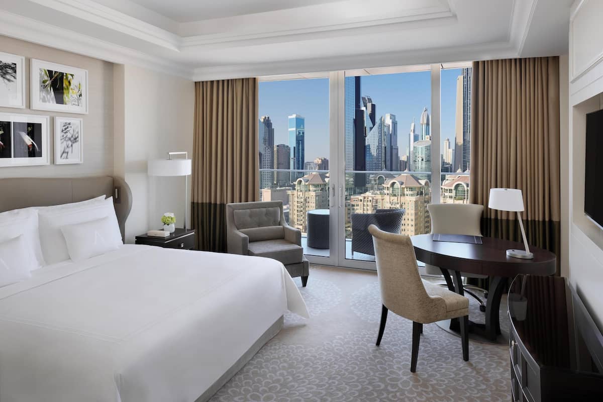 Quarto de hotel com uma cama grande, uma escrivaninha com cadeira, televisão e vista para o horizonte urbano através de grandes janelas.