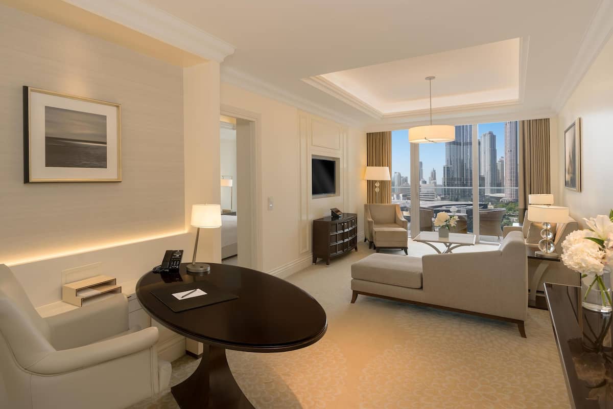 Quarto de hotel moderno com uma grande janela oferecendo vista da cidade, uma mesa de jantar redonda, um sofá e uma TV na parede.