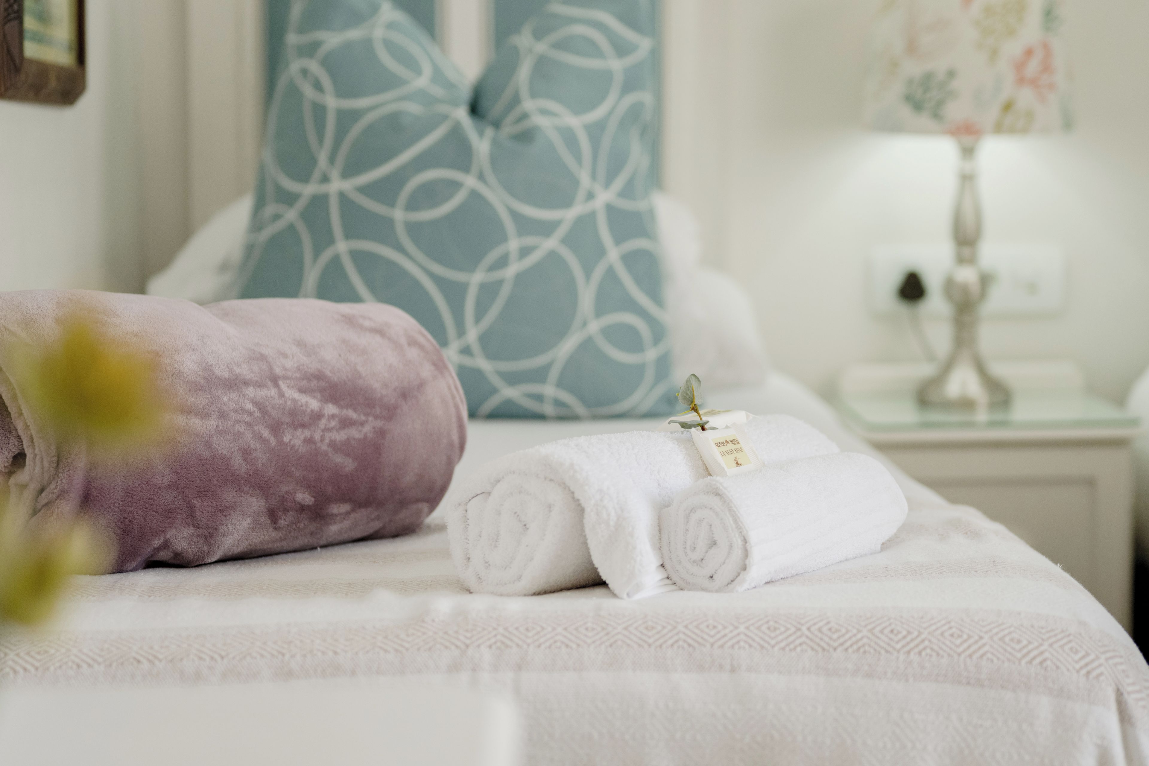 Uma cama com uma toalha branca enrolada e uma toalha roxa enrolada, uma mesinha de cabeceira branca com uma luminária, e um travesseiro azul com padrão branco.