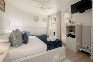 Quarto com cama, mesas de cabeceira, guarda-roupa, ventoinha no teto, janela com cortinas e televisão na parede.