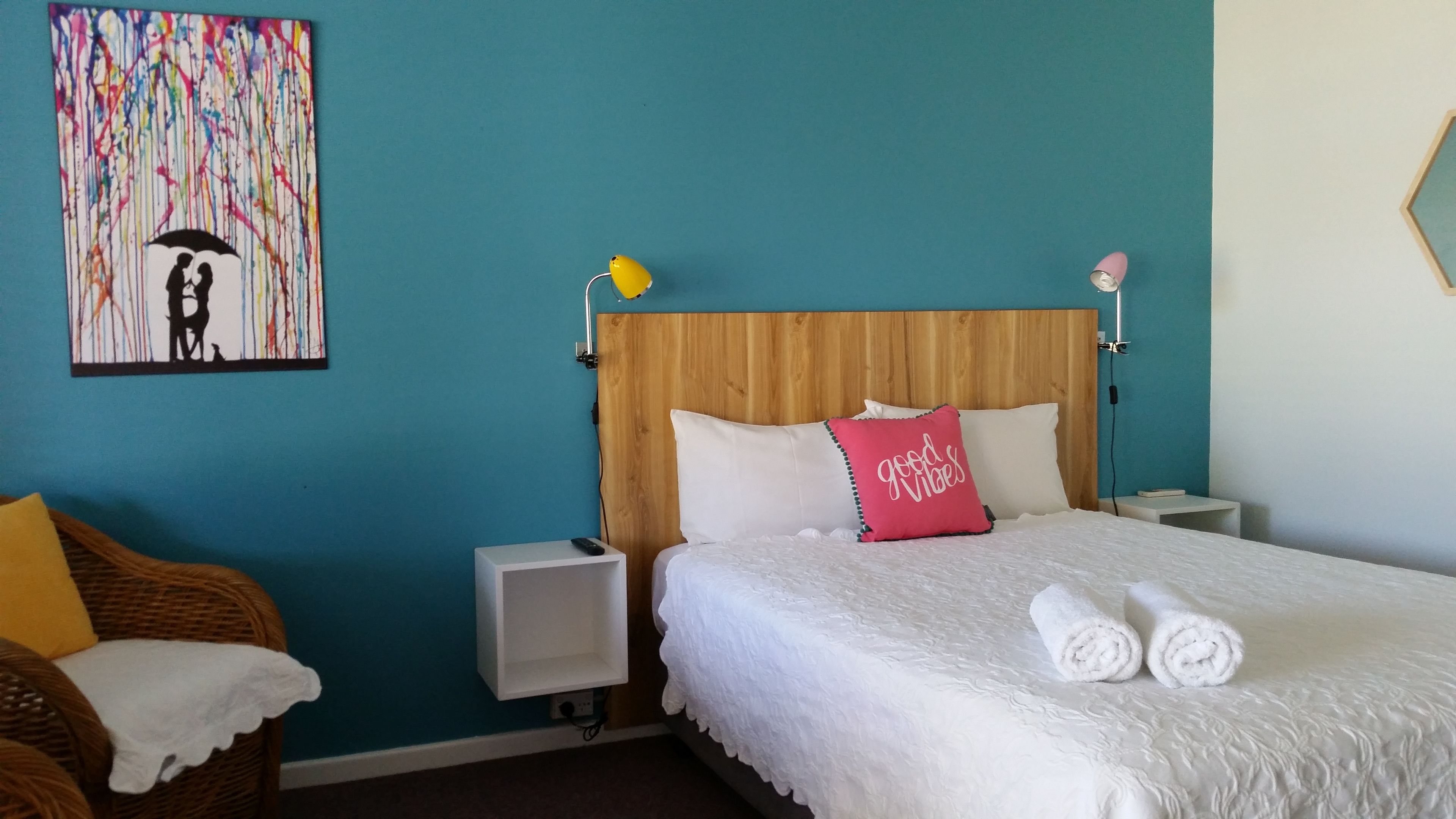 Quarto com cabeceira de madeira, roupa de cama branca, almofada rosa com a inscrição 'good vibes' e uma cadeira de vime com almofada amarela, além de um quadro abstrato e colorido na parede.