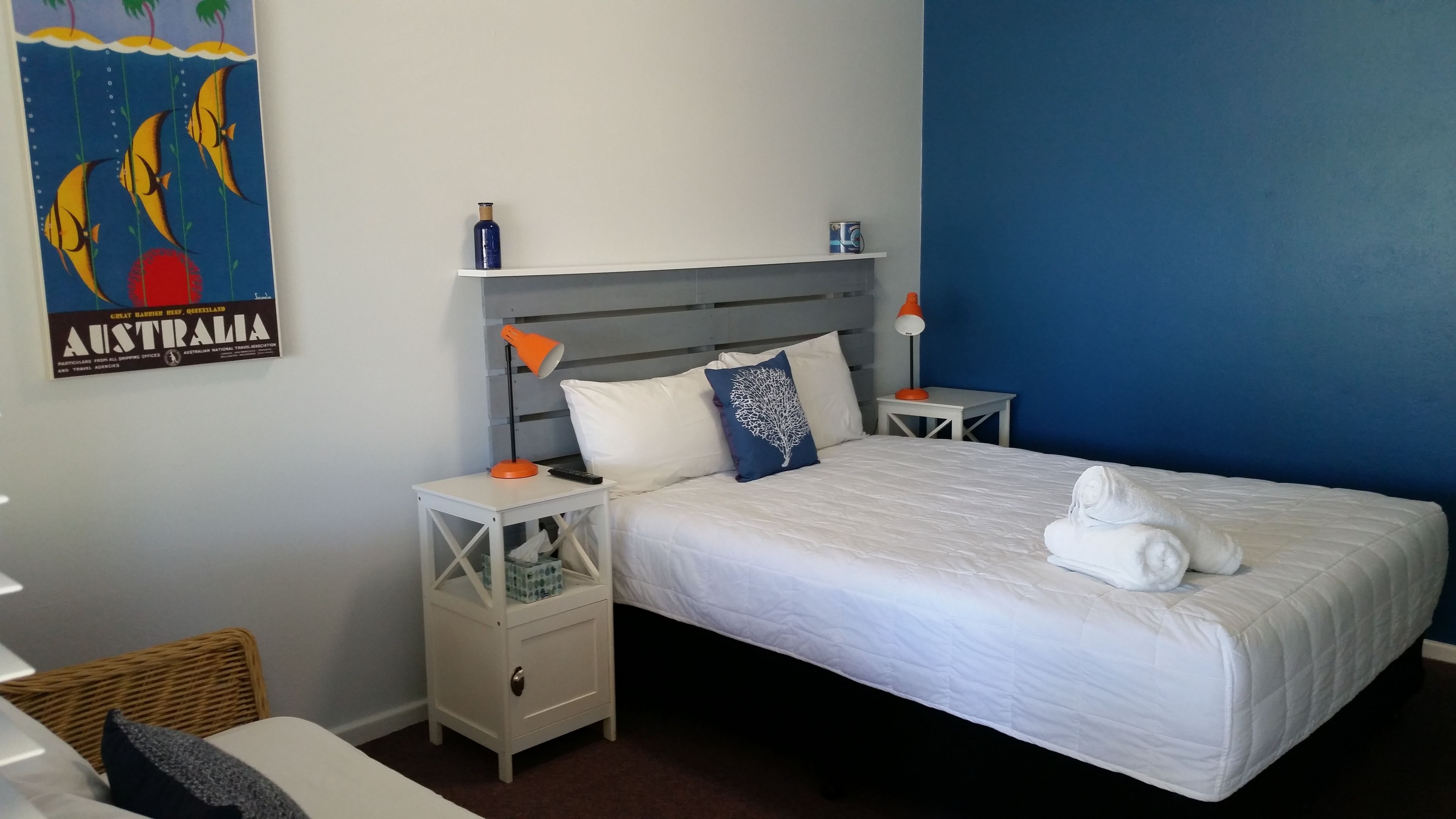 Quarto de hotel bem arrumado com uma cama, duas mesas de cabeceira, uma luminária e uma cadeira de vime, além de um quadro na parede.