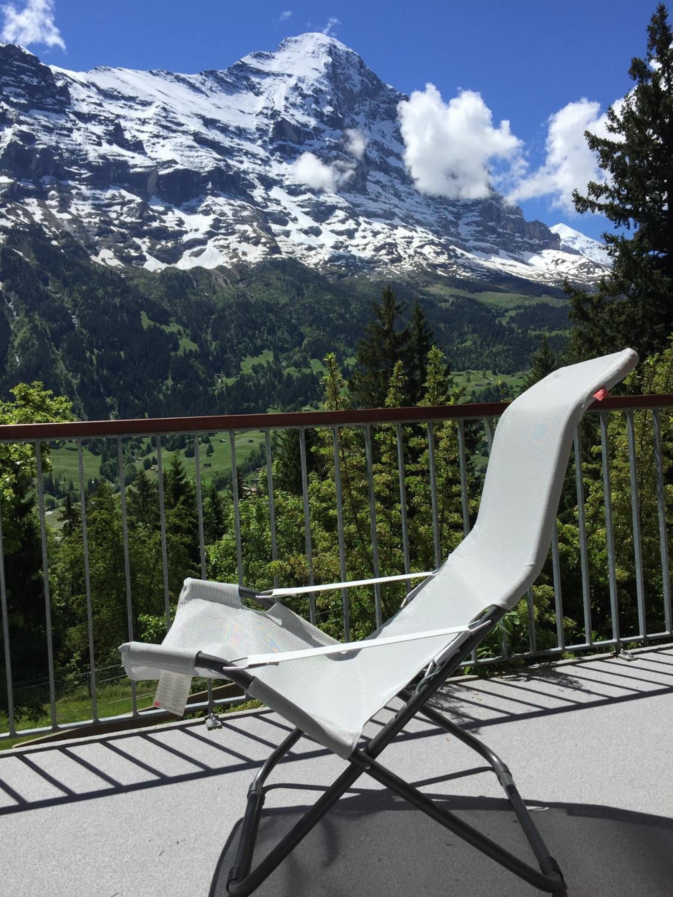 Uma cadeira de lounge branca numa varanda com guarda-corrimão de metal, com vista para uma paisagem montanhosa com picos nevados e vales verdes.