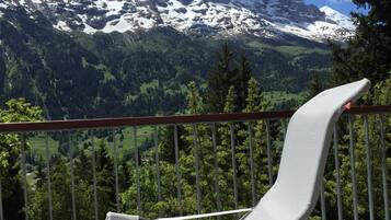 Uma cadeira de lounge branca numa varanda com guarda-corrimão de metal, com vista para uma paisagem montanhosa com picos nevados e vales verdes.