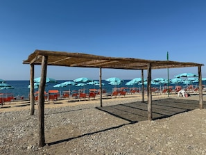 Uma praia com um pavilhão de madeira, cadeiras de praia vermelhas e guarda-sóis azuis.