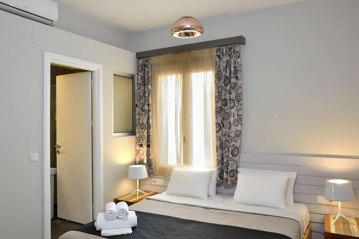 Quarto de hotel com cama, abajur, mesa de cabeceira, janela com cortinas estampadas e porta para um banheiro.
