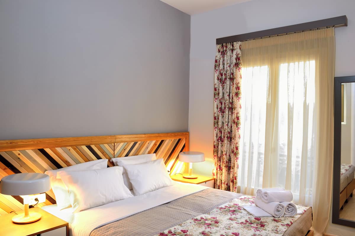 Uma cama bem arrumada com lençóis brancos, cabeceira de madeira e colcha estampada com flores. Há uma mesa de cabeceira com uma luminária e uma toalha dobrada.