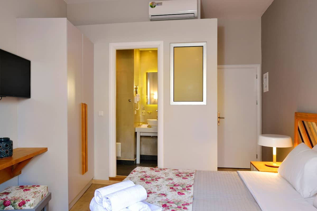 Quarto de hotel com cama, mesa de cabeceira com abajur, televisão na parede, banheiro com pia e espelho, e aparelho de ar condicionado.