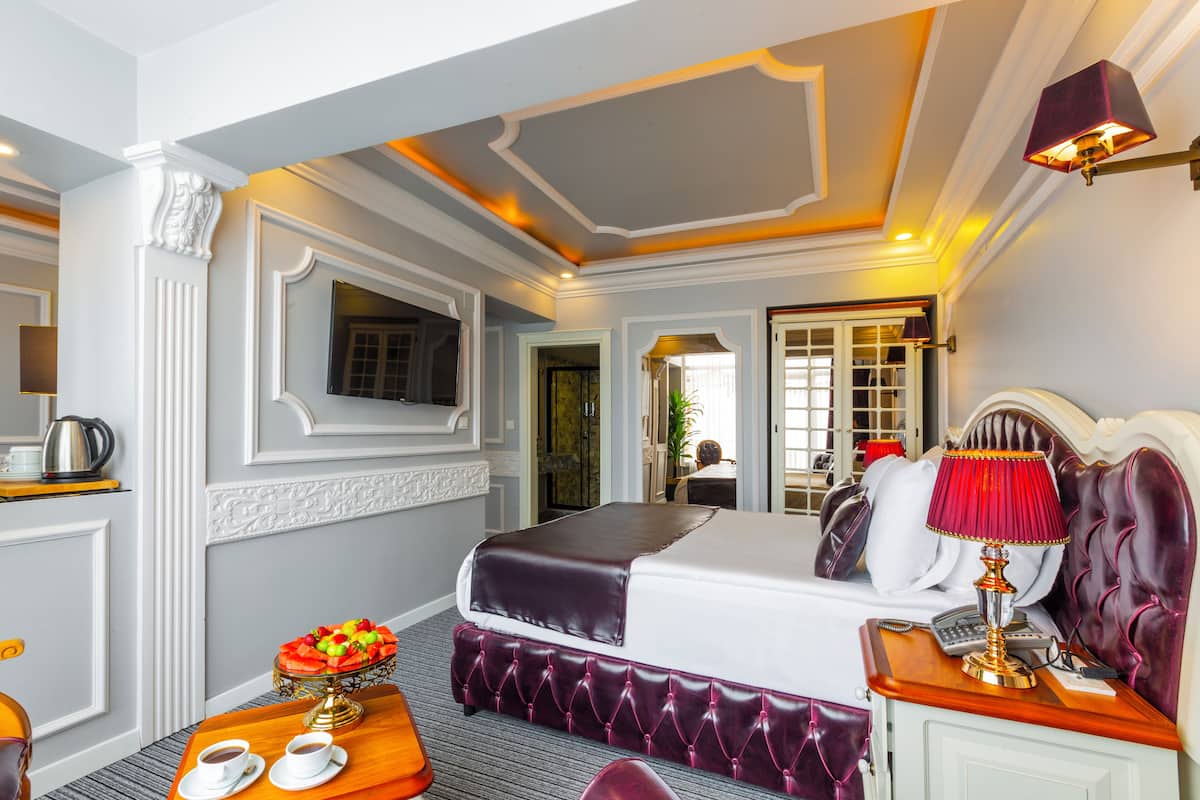 Quarto de hotel com uma cama grande, uma mesa de cabeceira com abajur, uma mesinha com uma cesta de frutas e chávenas, e um espelho na parede.