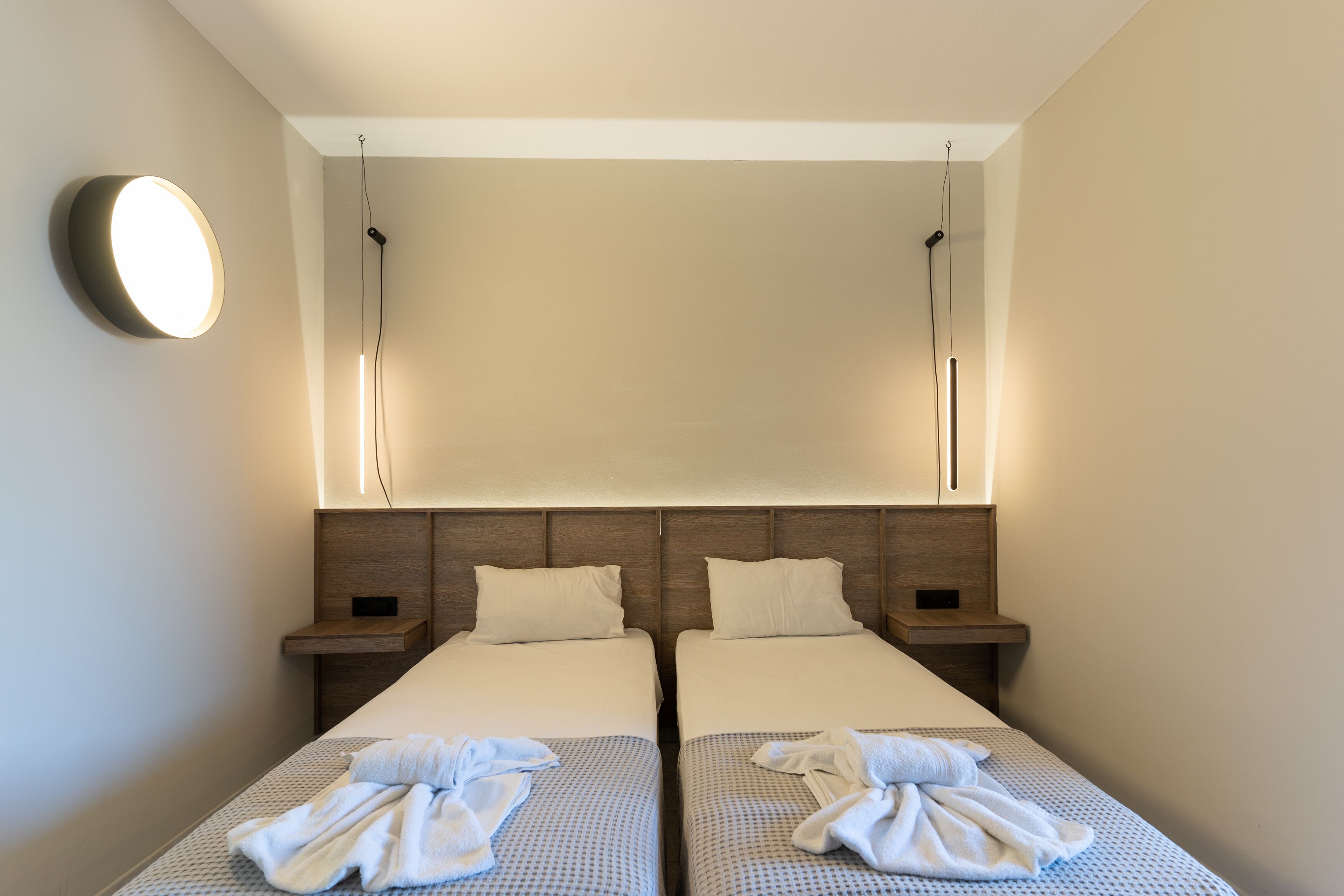 Quarto de hotel com duas camas, uma luminária redonda na parede e uma cabeceira de madeira.