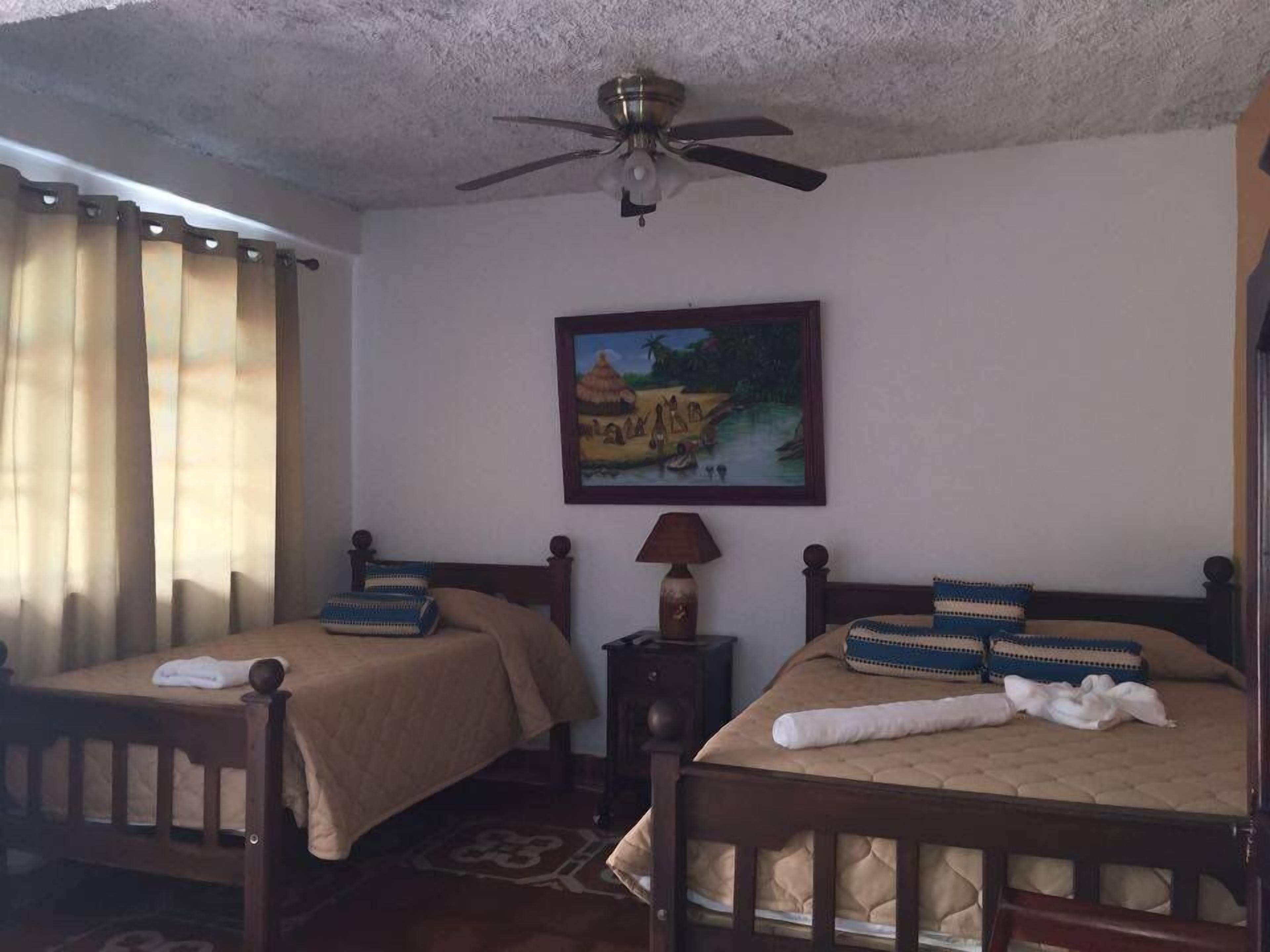 Quarto de hotel com duas camas, um ventilador de teto, um quadro e uma mesa de cabeceira.
