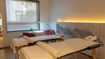 Um quarto com duas mesas de massagem, uma janela com persianas, uma escrivaninha com abajur e uma obra de arte pendurada na parede.