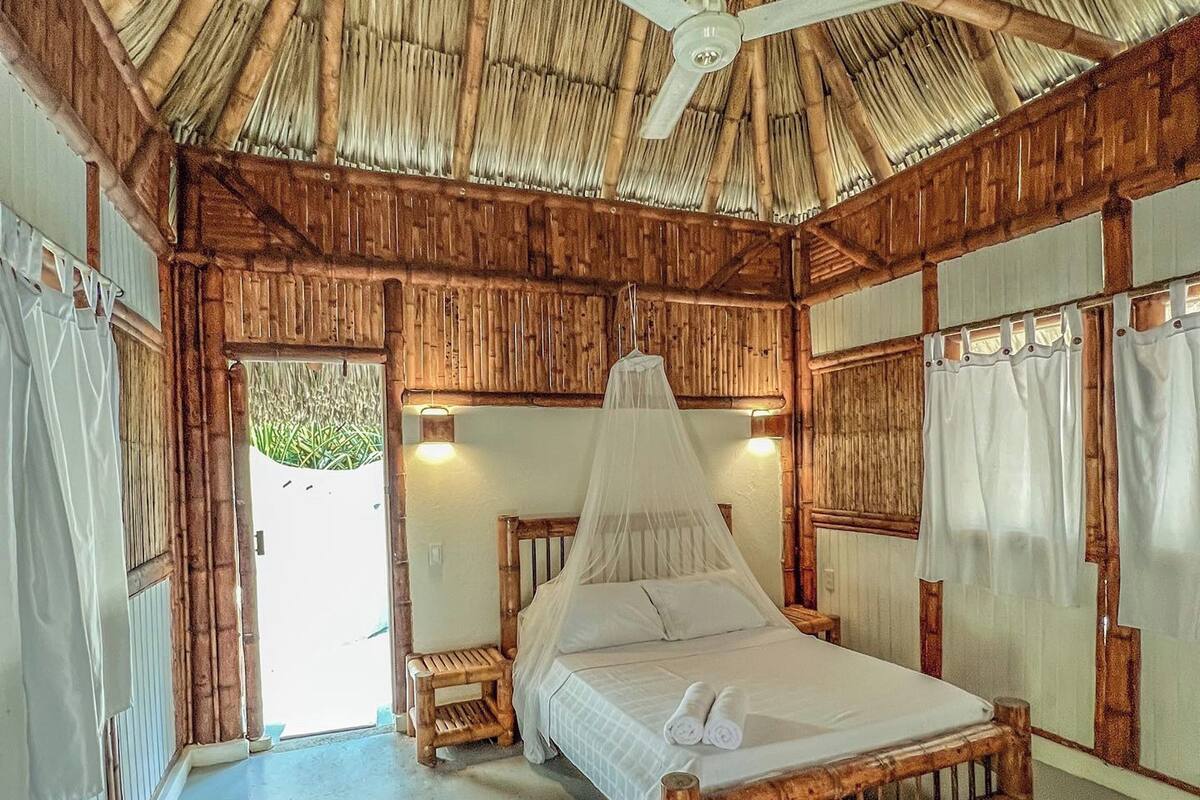 Quarto com uma cama, um banquinho de madeira, um ventilador de teto e uma rede mosquiteira.