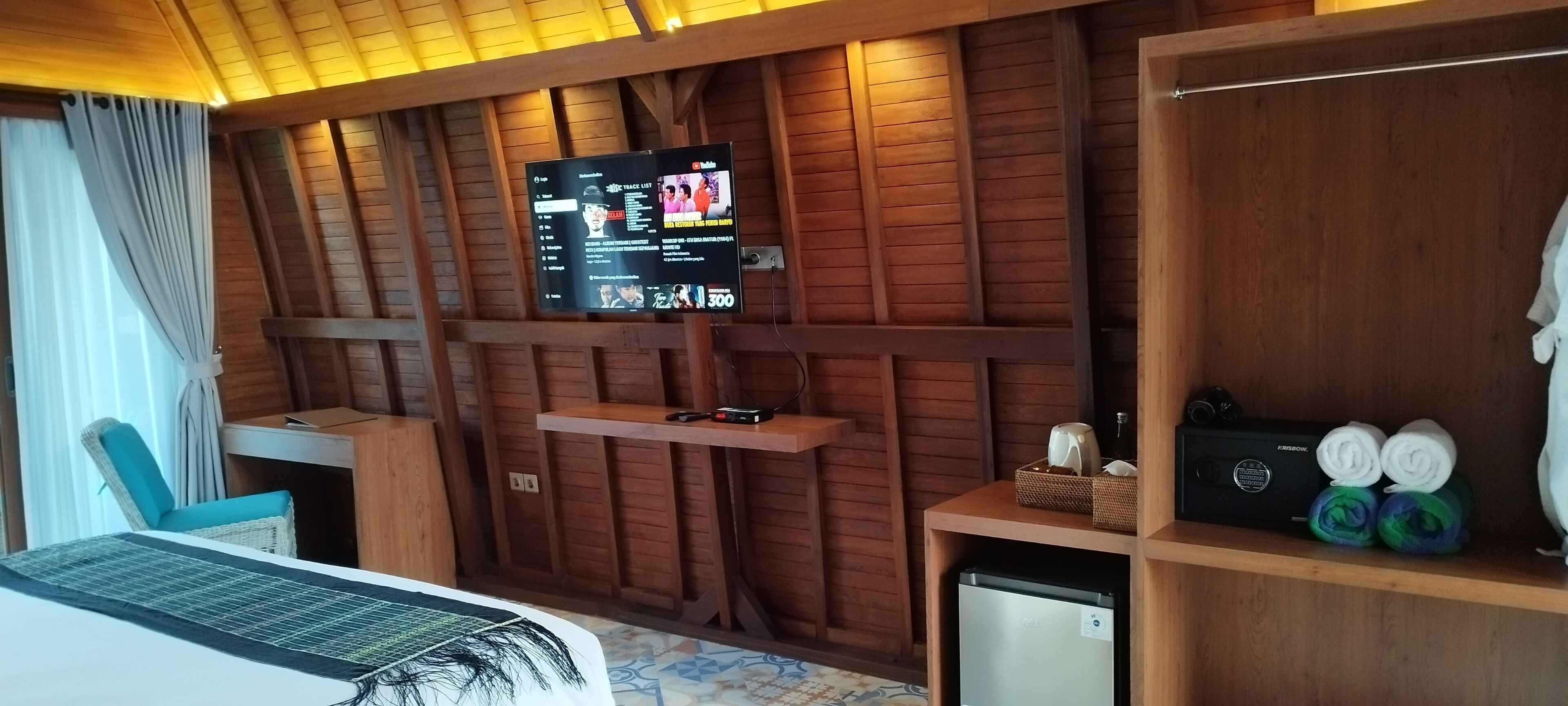 Quarto de hotel com parede de madeira, televisão fixada na parede, secretária com cadeira, mini-frigorífico e prateleira com toalhas.