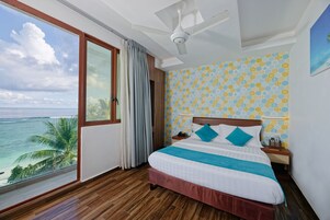 Quarto de hotel com uma grande janela com vista para o oceano, uma cama com roupa de cama azul e branca, um ventilador de teto e uma parede estampada.