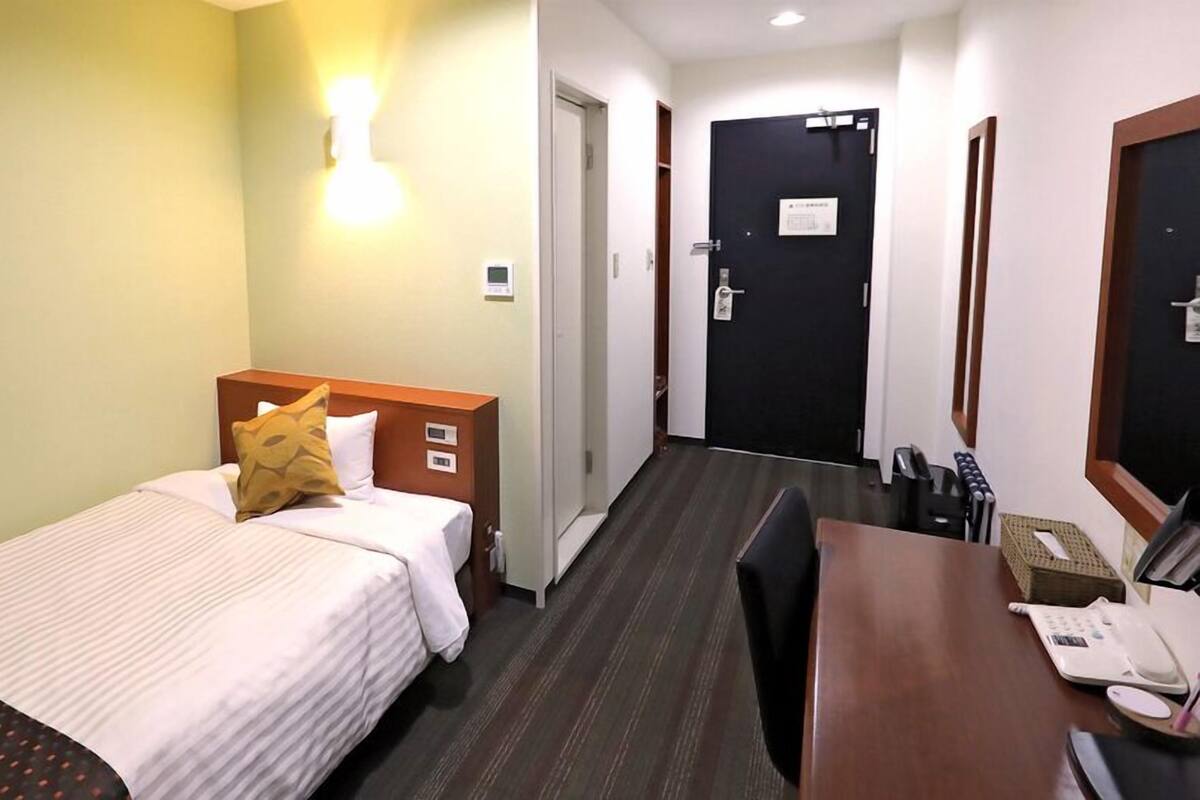 Quarto de hotel com cama, escrivaninha, cadeira, telefone e porta.