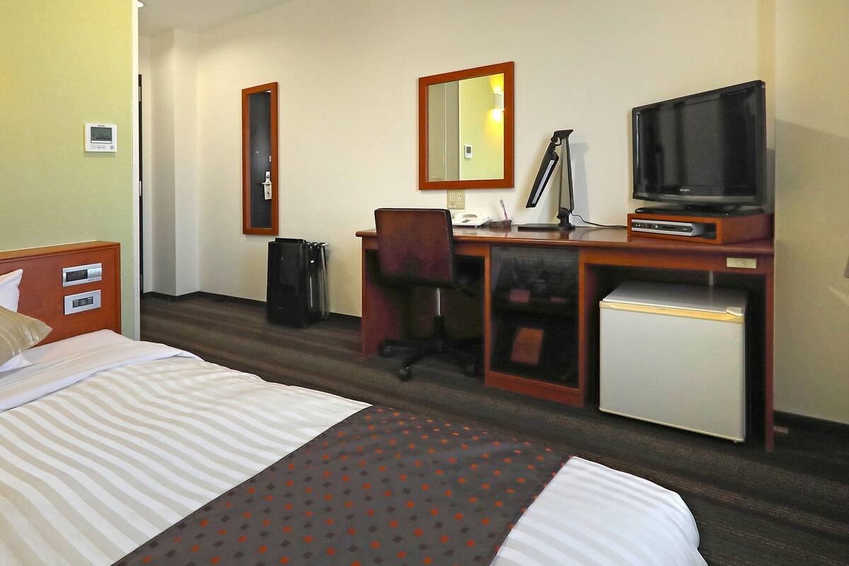 Quarto de hotel com cama, escrivaninha, cadeira, televisão e um pequeno frigorífico.