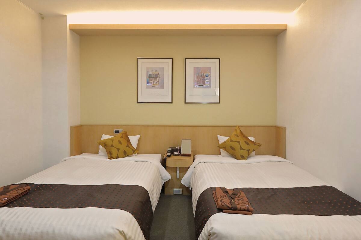 Quarto de hotel com duas camas de solteiro, uma mesa de cabeceira com luminária, um relógio e duas quadros na parede.