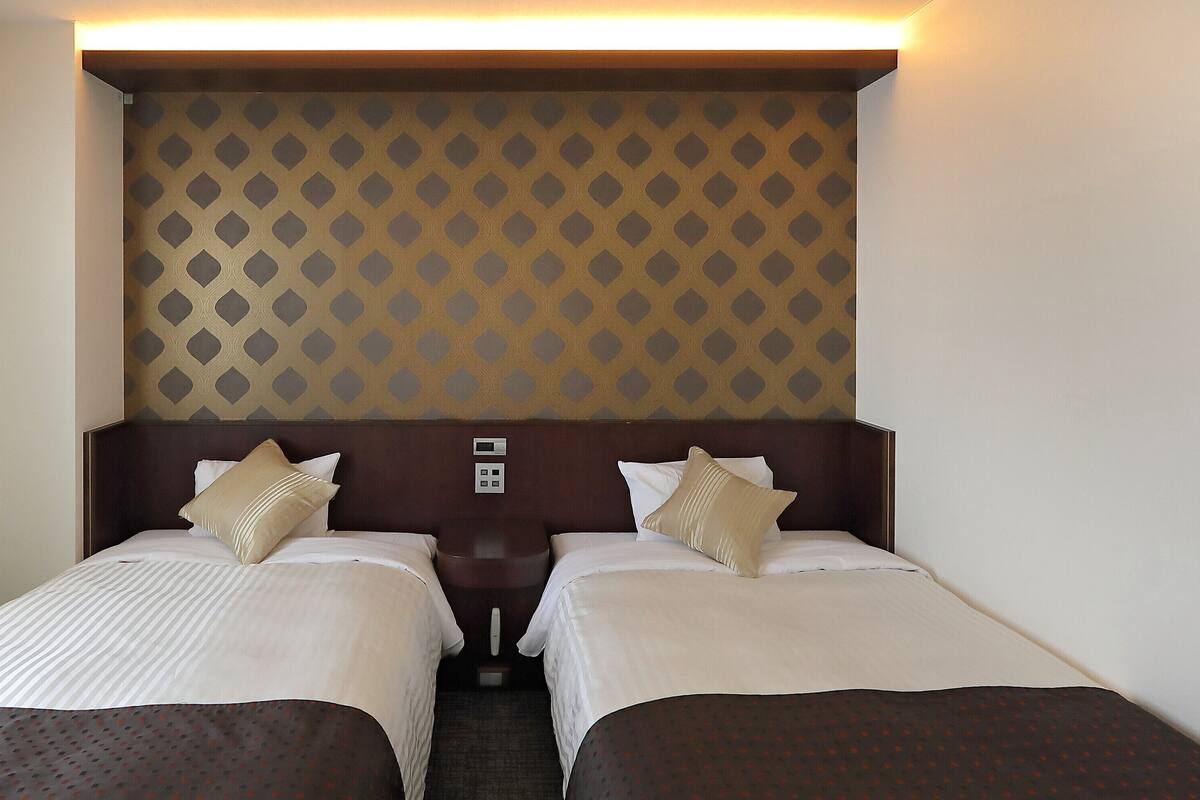 Quarto de hotel com duas camas de solteiro, cabeceira com padrão decorativo e painel de controle acima das camas.