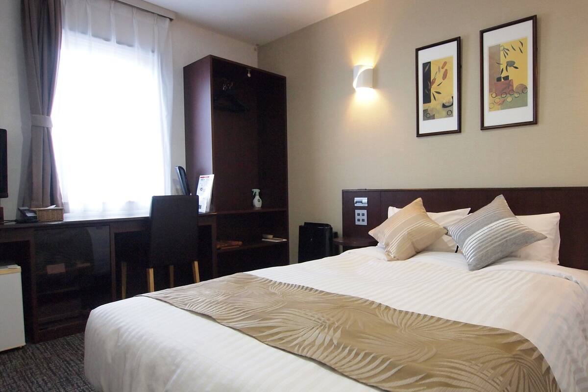 Quarto de hotel com uma cama grande, uma escrivaninha, uma cadeira e um guarda-roupa. Há duas luminárias fixadas na parede e duas obras de arte emolduradas sobre a cama.