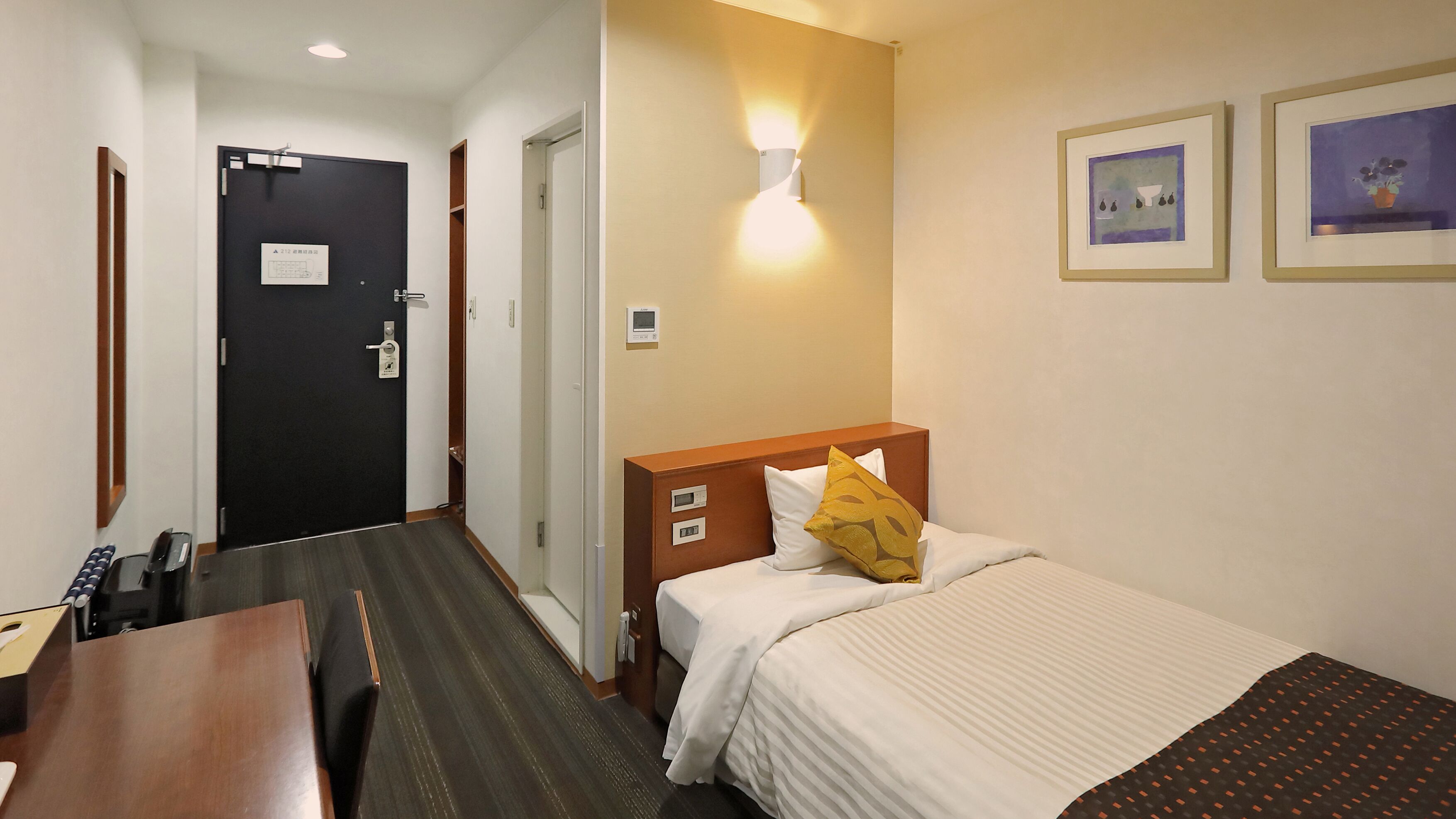 Quarto de hotel com cama, escrivaninha, cadeira, luminária na parede e duas imagens emolduradas.