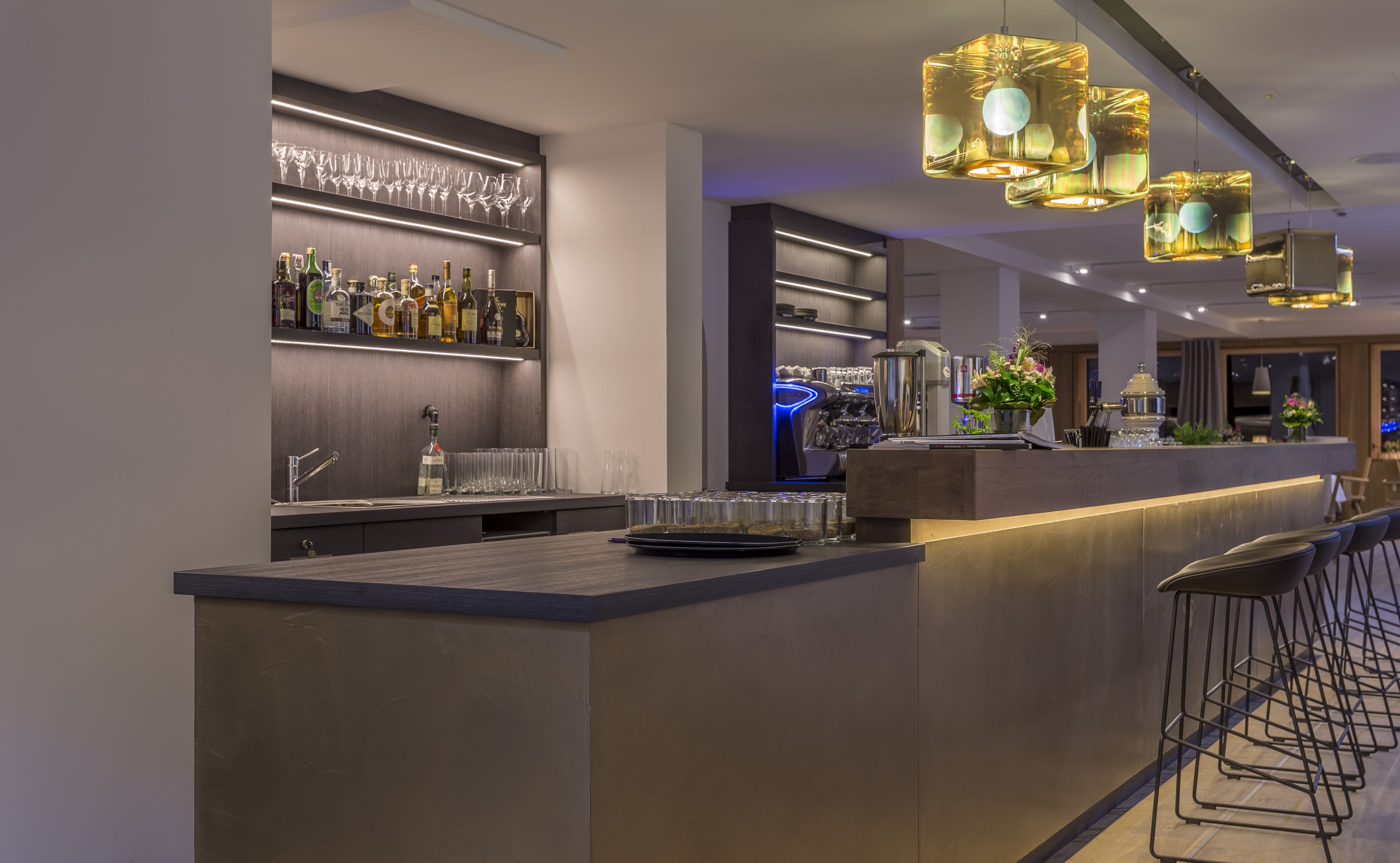 Uma área de bar moderna com ilha central, banquetas, prateleiras com garrafas e iluminação neon no fundo.