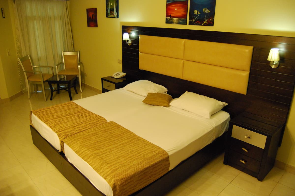 Quarto de hotel com uma cama grande, uma mesa de cabeceira com abajur, uma cadeira e um criado-mudo com telefone.