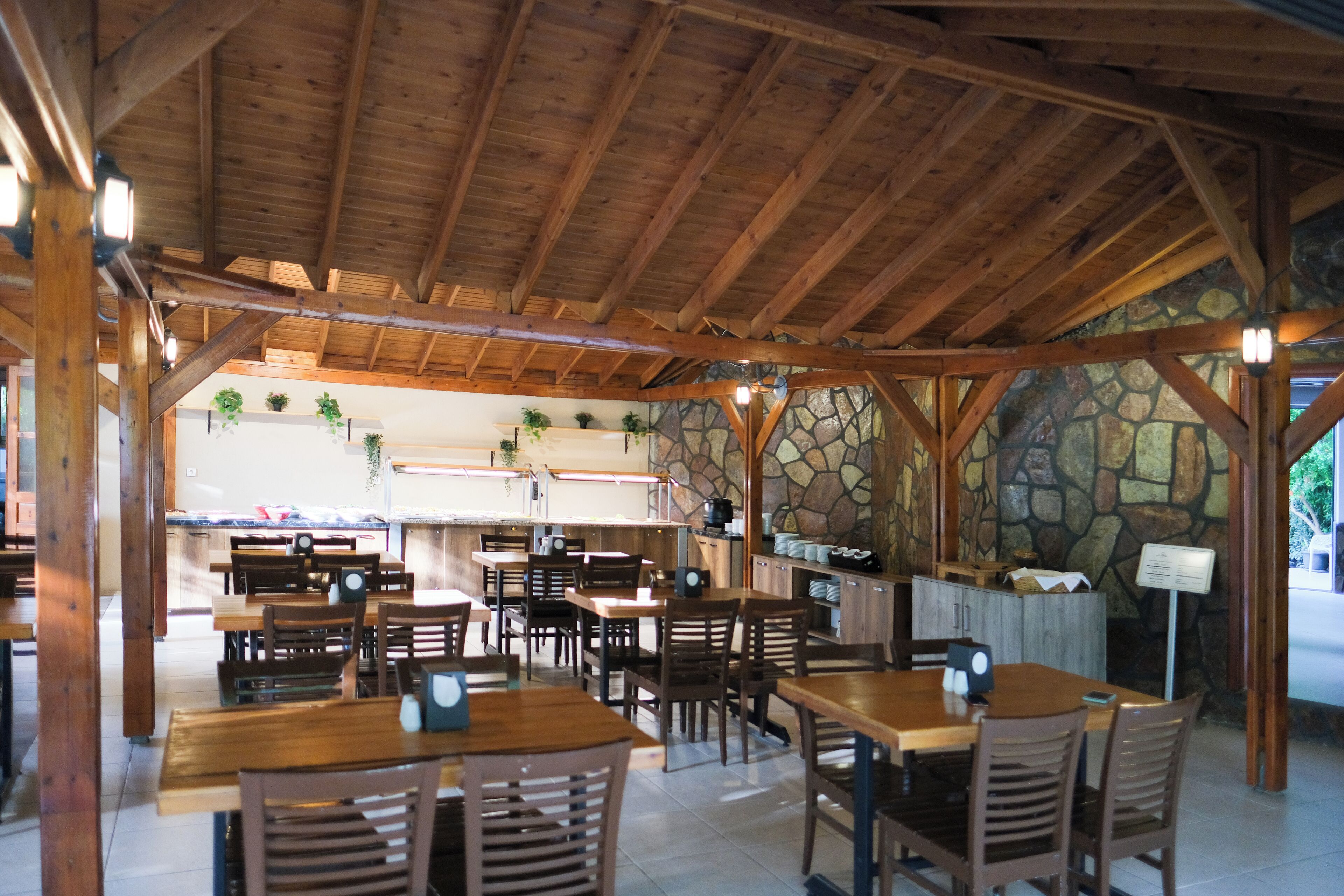 Um restaurante com mesas e cadeiras de madeira, parede de pedra e área de cozinha ao fundo.