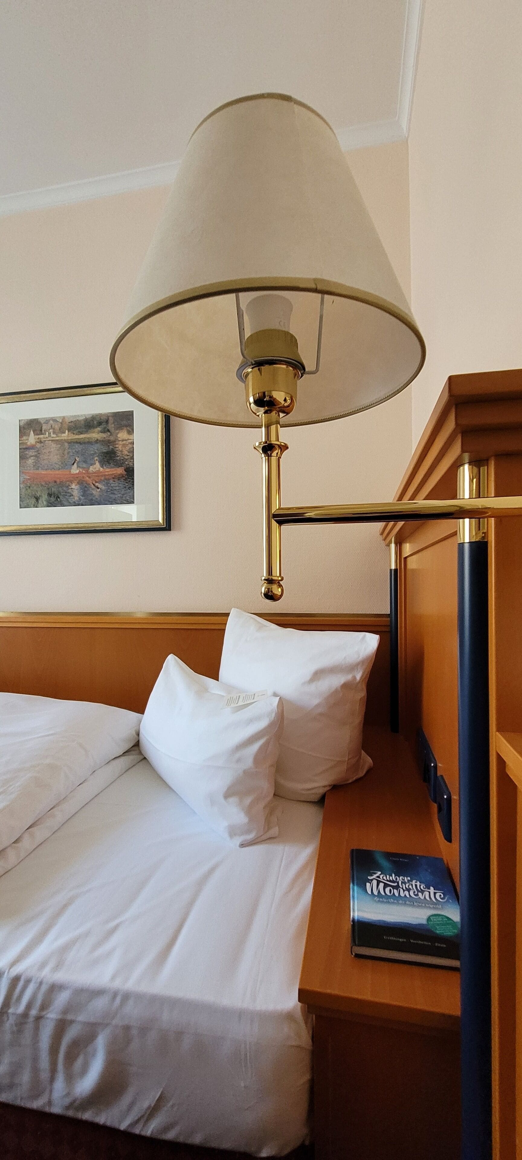 Quarto de hotel com uma cama bem arrumada, abajur, quadro na parede e um livro na mesinha de cabeceira.