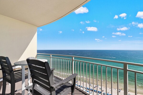 Varanda com duas cadeiras e uma mesa, com vista para uma praia de águas azuis cristalinas e algumas pessoas.