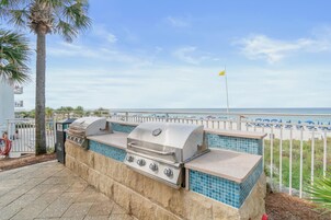 Churrasqueira com painel de azulejos, uma palmeira e vista para a praia com guarda-sóis.
