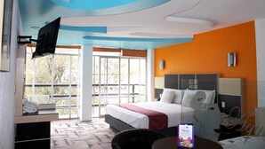 Quarto de hotel moderno com uma cama grande, televisão de tela plana na parede, mesa de jantar e vista para o exterior por grandes janelas.