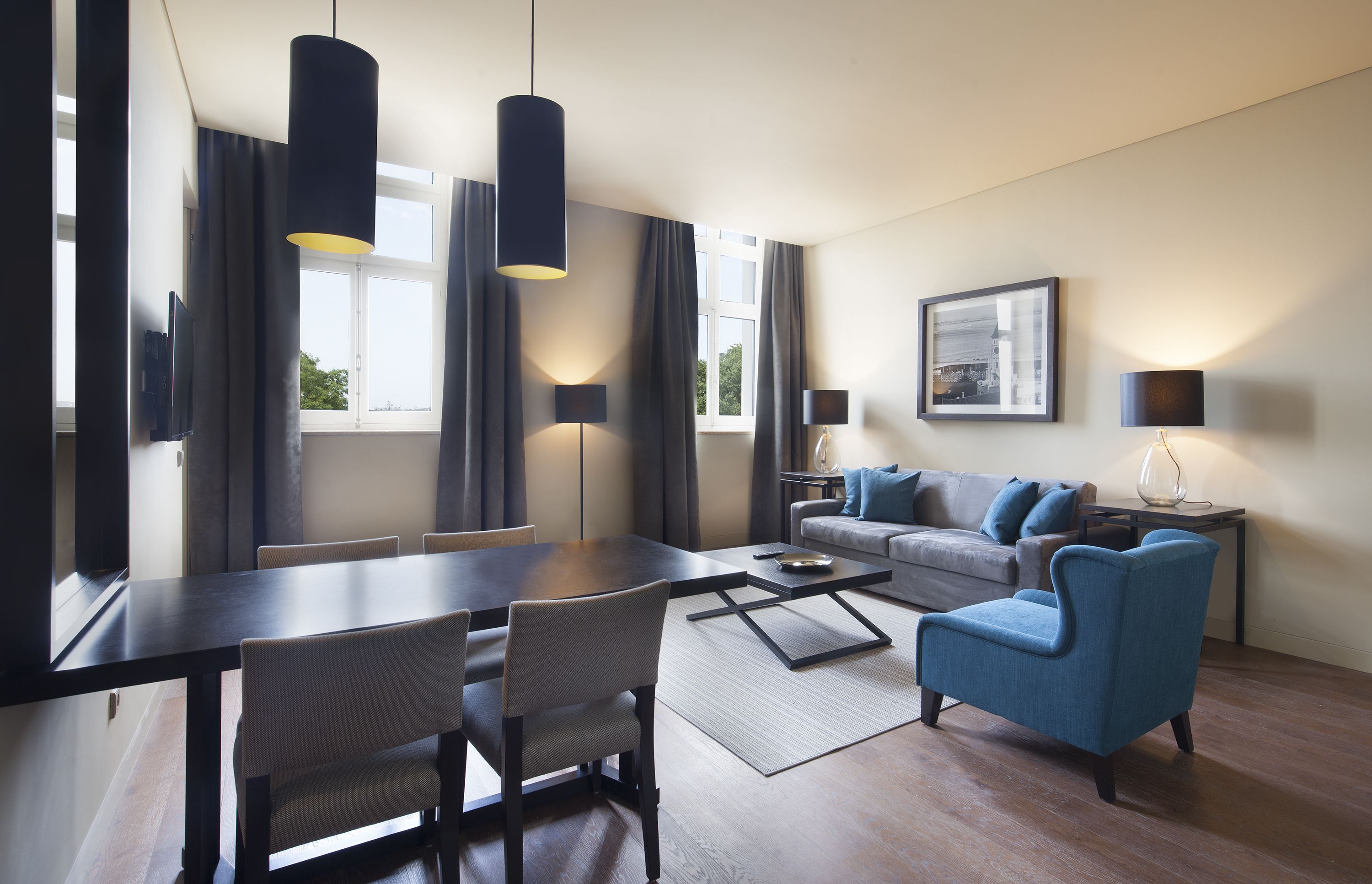 Uma sala de estar moderna com uma mesa de jantar de madeira escura, cadeiras, uma poltrona azul, um sofá e uma mesa de centro. Há duas luminárias de chão e um quadro na parede.