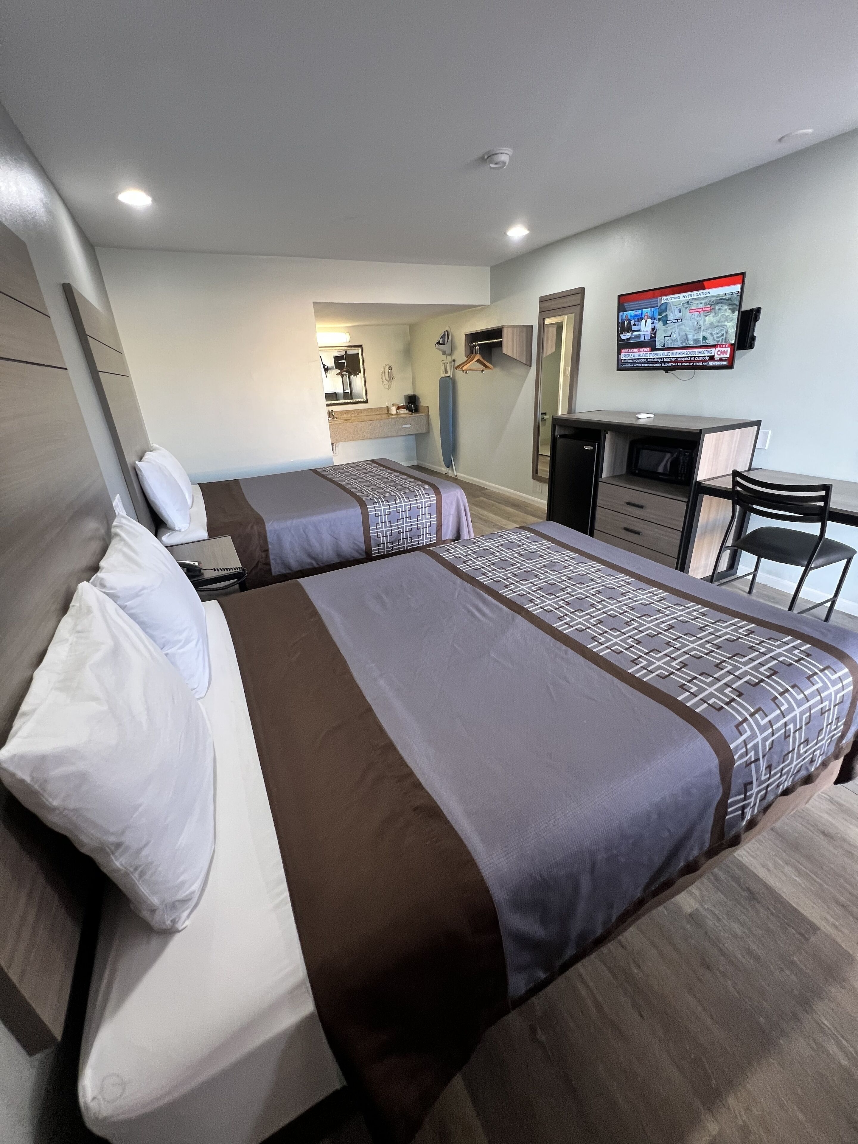 Quarto de hotel com duas camas, uma escrivaninha, uma cadeira, televisão presa à parede e uma pequena cozinha equipada.