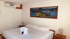 Uma cama bem arrumada com lençóis brancos, um quadro de uma paisagem costeira na parede e um ar condicionado Samsung montado acima da cama.