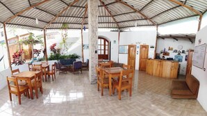 Um quarto espaçoso com teto alto e cobertura de metal corrugado. Possui piso de madeira, várias mesas e cadeiras de madeira e um pequeno balcão de recepção.
